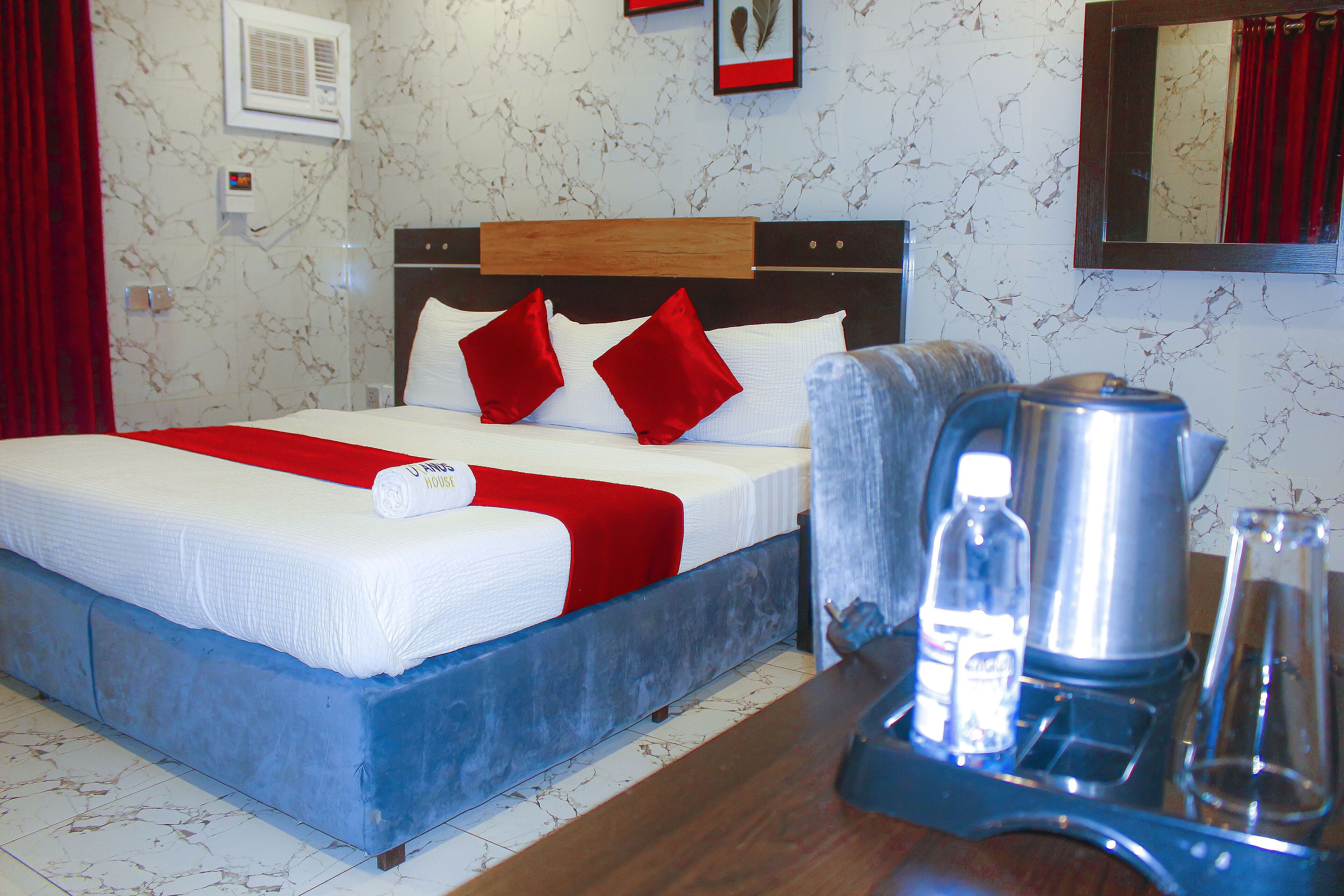 Quarto de hotel com cama, cabeceira de madeira, colcha vermelha e branca, e mesa de cabeceira com chaleira e garrafa.