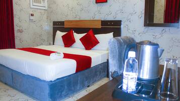 Quarto de hotel com cama, cabeceira de madeira, colcha vermelha e branca, e mesa de cabeceira com chaleira e garrafa.