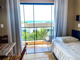 Quarto de hotel com uma cama, uma mesa de madeira e uma cadeira. H á uma janela grande com cortinas, oferecendo vista para o oceano e edifícios.