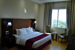 Quarto de hotel com uma cama grande, duas luminárias de cabeceira, uma mesinha, uma cadeira e vista para a cidade pela janela.