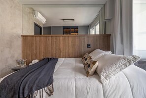 Quarto de hotel moderno com uma cama grande, cabeceira de madeira, mesa de cabeceira com abajur e um espelho amplo.