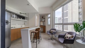 Uma cozinha moderna com ilha central, banquetas e um espaço de estar com duas cadeiras e uma mesa.
