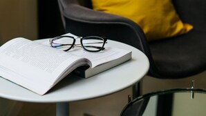 Uma cadeira com um travesseiro amarelo, uma mesa com um livro e óculos, e uma luminária ao fundo.