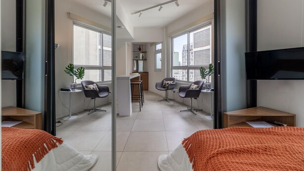 Quarto de hotel moderno com divisória de vidro, duas camas com mantas laranja, uma escrivaninha com cadeira, televisão e amplas janelas com vista para a cidade.