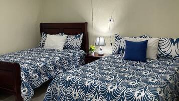 Quarto de hotel com duas camas, cada uma com colcha e almofadas estampadas. Há uma mesa de cabeceira com uma luminária e uma pequena planta.