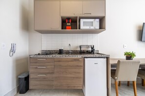 Cozinha compacta com micro-ondas, mini-geladeira e pia. Há também uma lixeira e uma cadeira à vista.