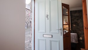 Uma porta azul clara com puxador prateado e caixa de correio, dando acesso a um quarto com parede de pedra e cama com cobertor branco.