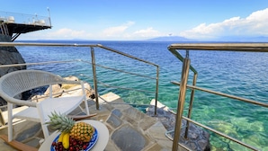 Um terraço de pedra com grade de metal, vista para águas azuis e cristalinas, uma cadeira de vime e uma mesa com frutas.