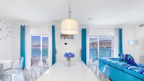 Uma área de refeições moderna com uma mesa branca, cadeiras transparentes e vista para o mar através de grandes janelas.