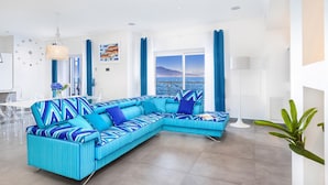 Uma sala de estar moderna com um amplo sofá em formato de L, uma área de refeições com cadeiras transparentes e vista para o mar através de grandes janelas.