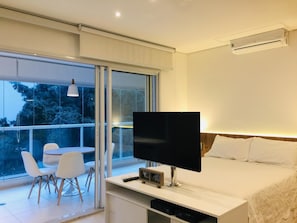 Quarto de hotel moderno com uma cama grande, televisão de tela plana, área de refeições com cadeiras e uma varanda com vista para árvores.