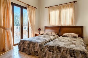 Quarto com duas camas, cabeceira de madeira, mesa de cabeceira com abajur e vista para uma varanda com cortinas.