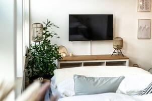 Quarto moderno com uma cama grande, televisão na parede, um banquinho de madeira e uma planta em vaso.