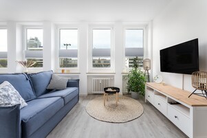 Uma sala de estar moderna com um sofá azul, uma mesa de centro redonda de madeira, uma TV sobre um suporte branco e janelas amplas com vista para a cidade.