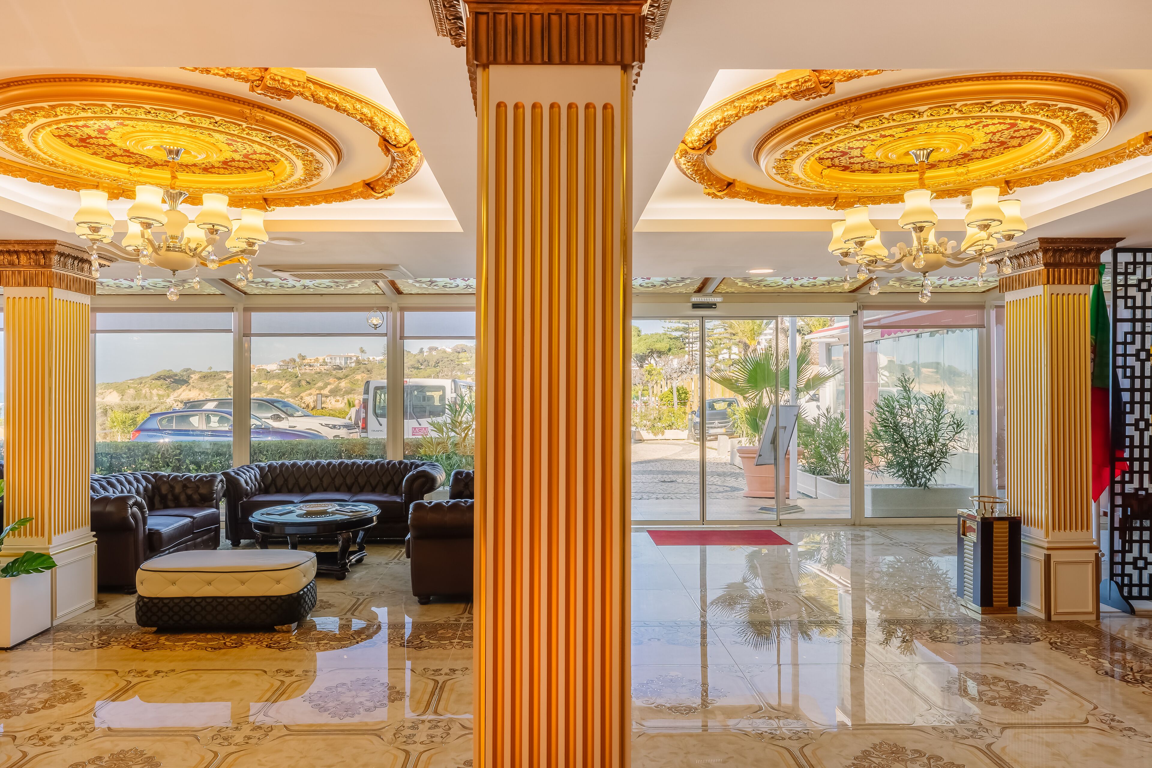 Um hall luxuoso com designs ornamentados no teto, uma área de estar com sofás de couro e vista para o exterior.