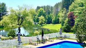 Piscina cercada por vegetação exuberante e um lago, com cadeiras de descanso e guarda-sóis, e uma cerca branca.