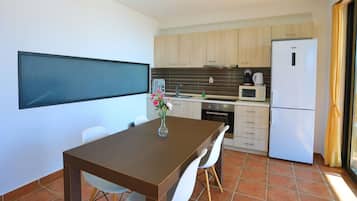 Uma cozinha moderna com área de refeições, apresentando uma televisão de tela plana grande, mesa de jantar de madeira, cadeiras brancas e uma geladeira.
