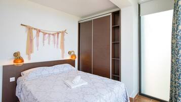 Uma cama bem arrumada com um cobre-leito estampado, um guarda-roupa de madeira e uma janela grande com cortina.