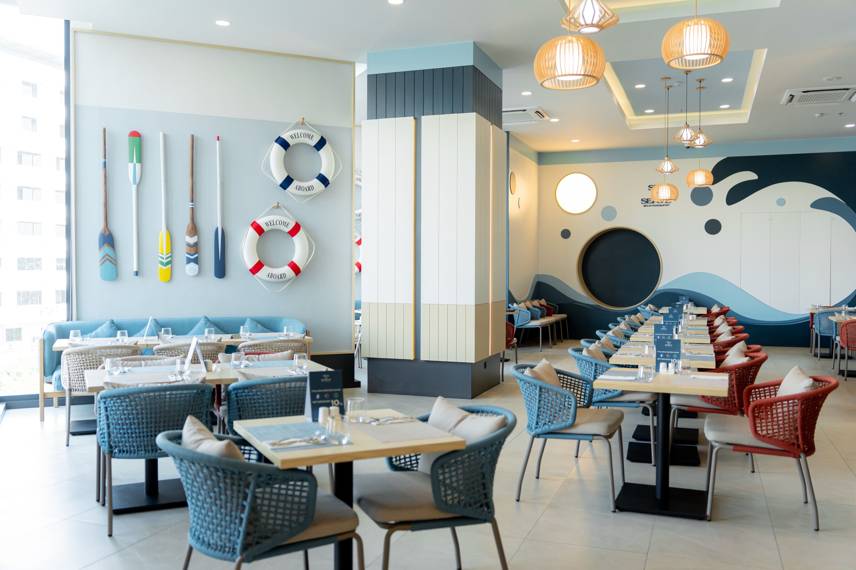 Um restaurante com cadeiras de vime azul e vermelha, mesas brancas e decoração náutica, incluindo boias salva-vidas e remos.