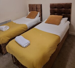 Duas camas de solteiro com cabeceiras castanhas e roupa de cama branca, cada uma com um cobertor amarelo e uma toalha branca enrolada.