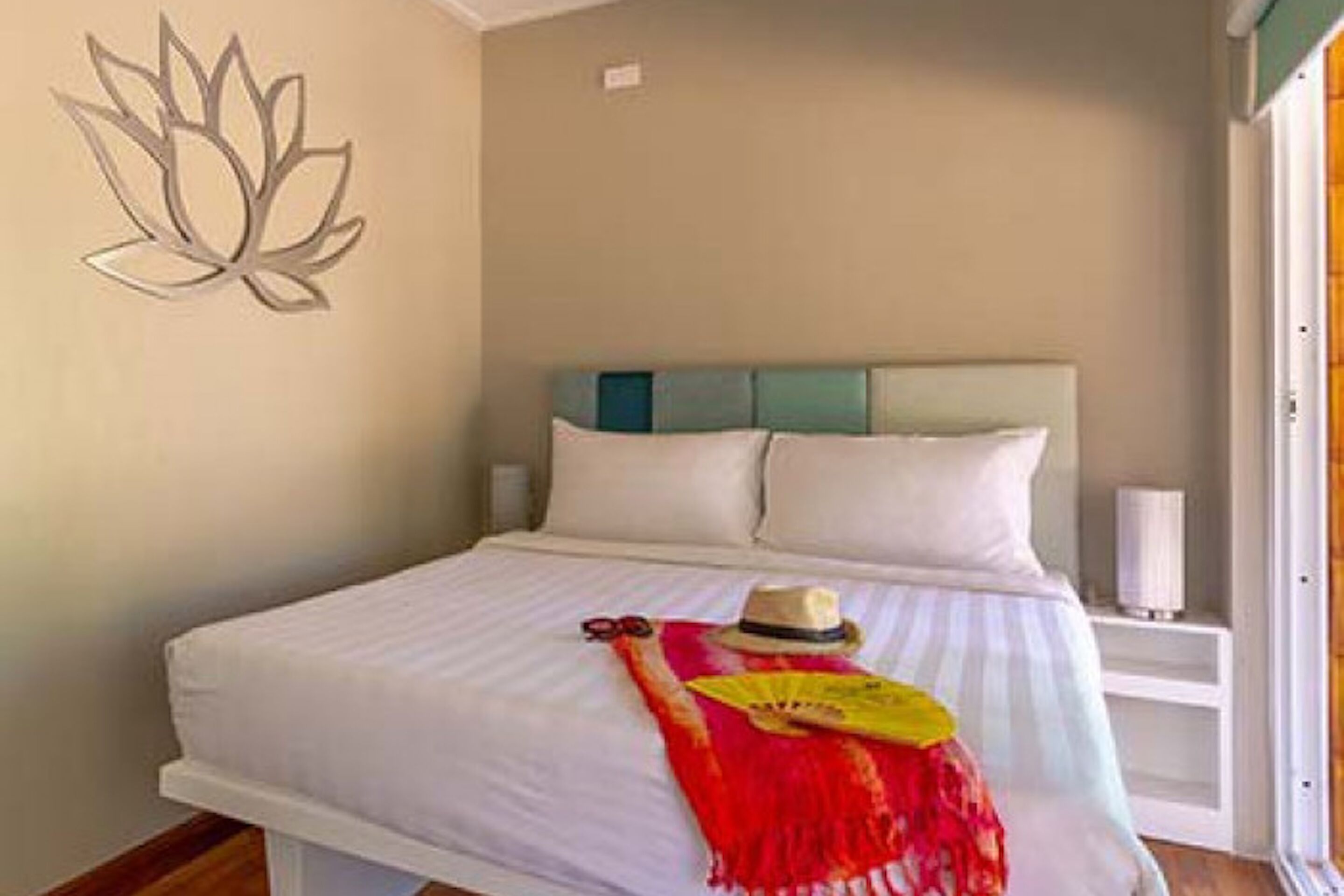 Cama bem arrumada com edredom branco, manta vermelha e amarela, e um chapéu em cima.