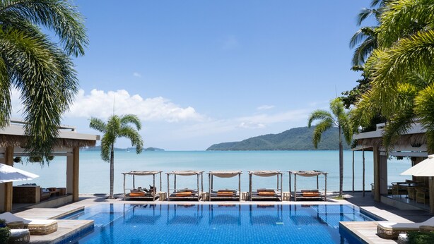 Área na piscina com espreguiçadeiras e guarda-sóis, com vista para o mar azul e colinas ao longe.