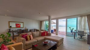 Uma sala de estar espaçosa com um sofá grande, uma mesa de centro e uma área de jantar. Há uma janela ampla com vista para o mar e uma varanda com móveis de exterior.