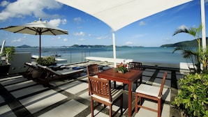 Um terraço com mesa e cadeiras de madeira, um guarda-sol branco e vista para o mar.