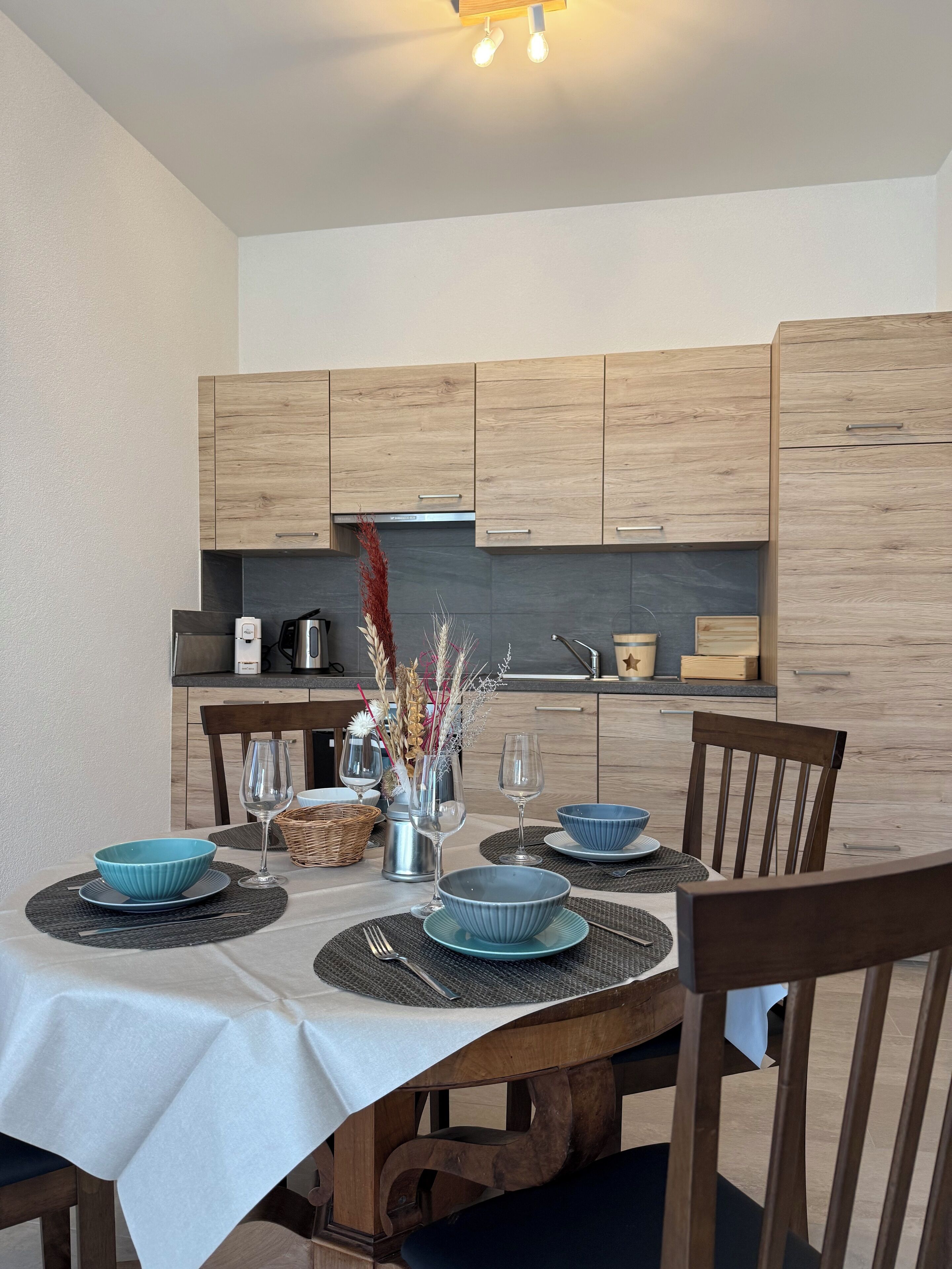 Uma sala de jantar com uma mesa posta para quatro pessoas, rodeada por cadeiras de madeira. Uma cozinha com armários de madeira e um fogão moderno é visível ao fundo.