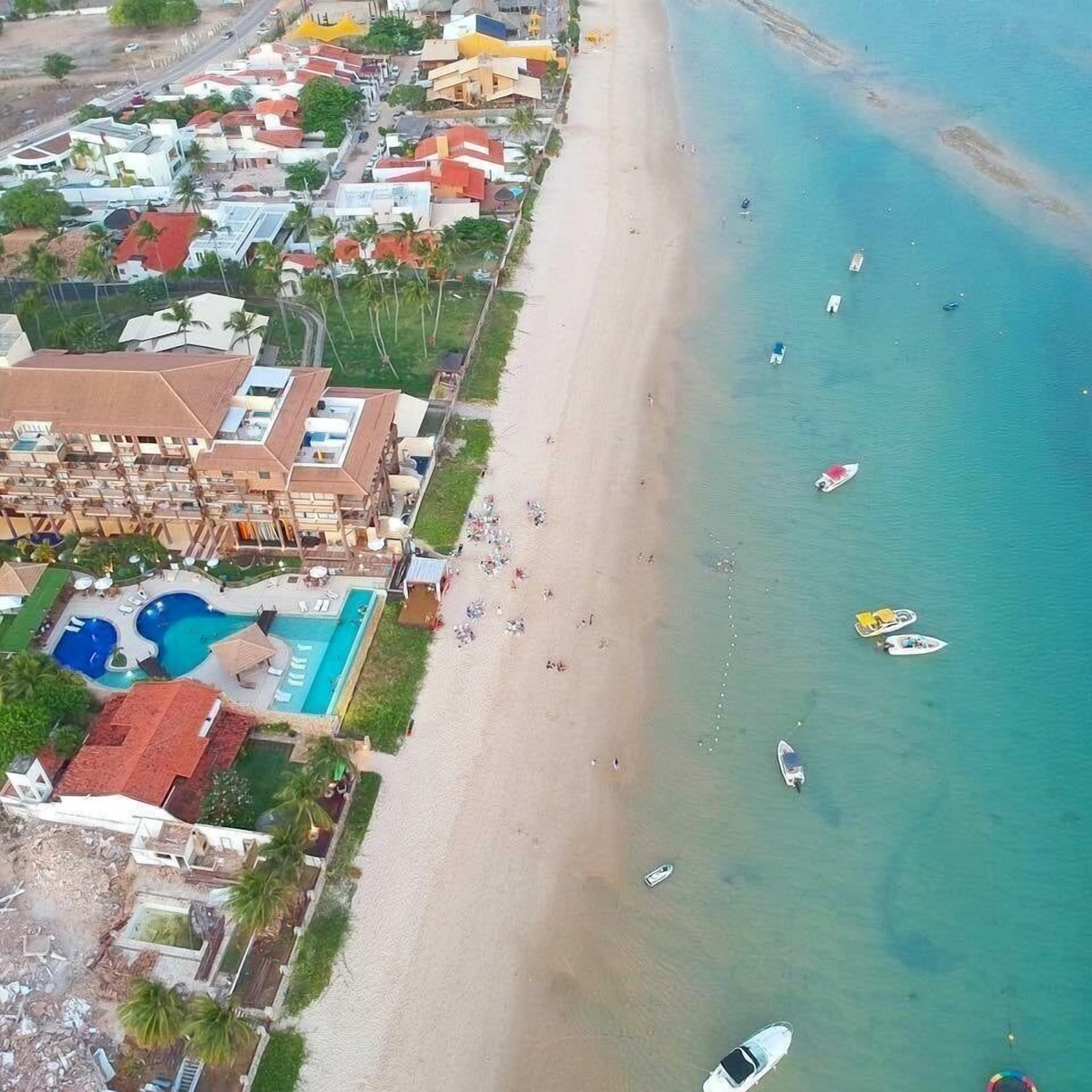 Vista aérea de um resort costeiro com uma grande piscina, praia e vários barcos na água.