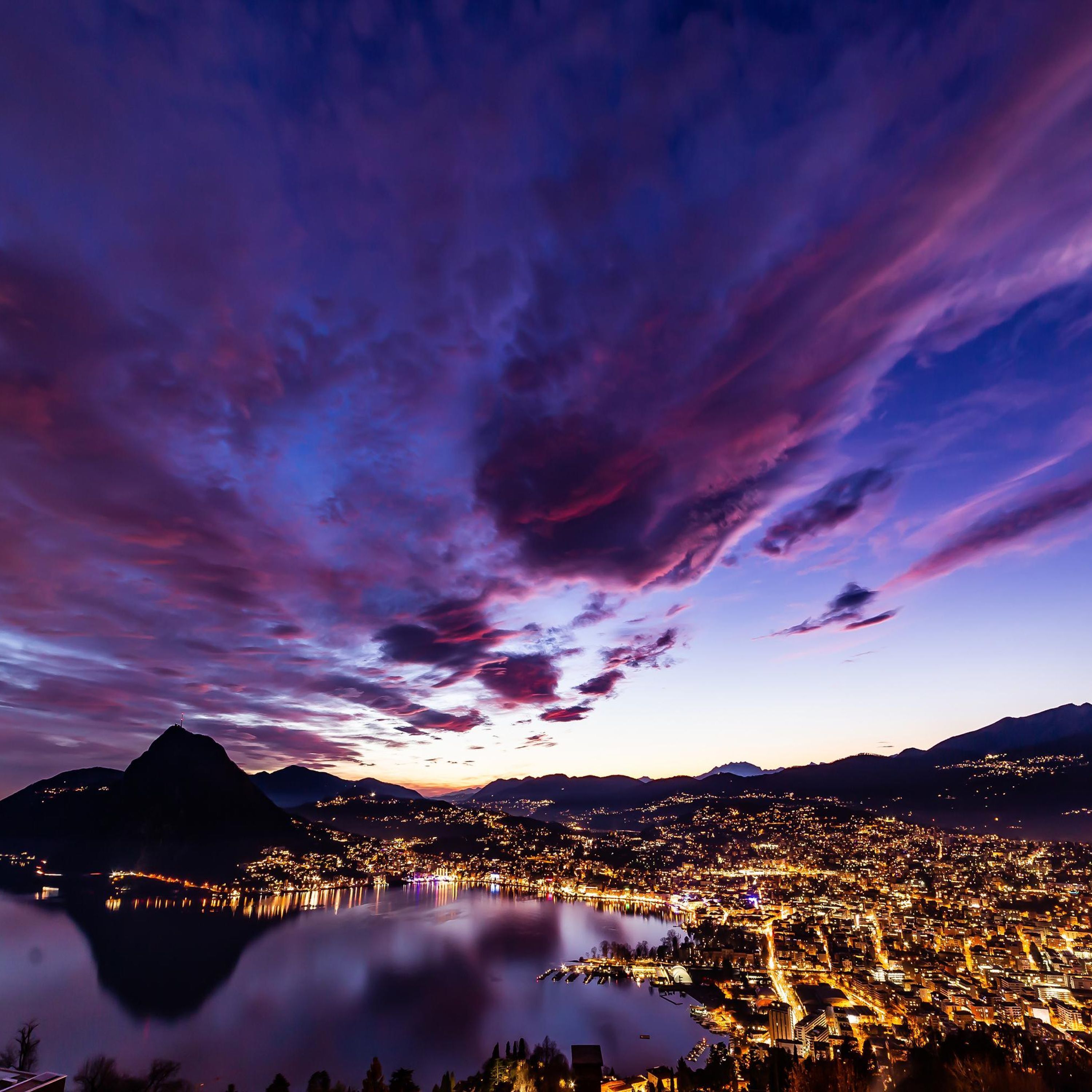Vista panorâmica de uma cidade ao entardecer, com uma montanha imponente em primeiro plano e um corpo d'água refletindo as luzes da cidade.