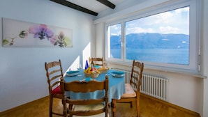 Uma sala de jantar com uma mesa posta para quatro pessoas, uma janela com vista para um lago e montanhas, e um quadro de flores na parede.
