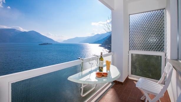 Uma varanda com mesa de vidro, uma garrafa e copos, com vista para um corpo d'água e montanhas.