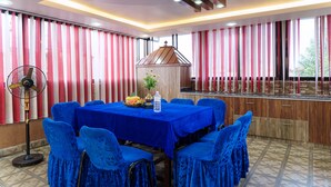Uma sala de jantar com toalha de mesa azul, cadeiras azuis, um ventilador e uma cesta de frutas.