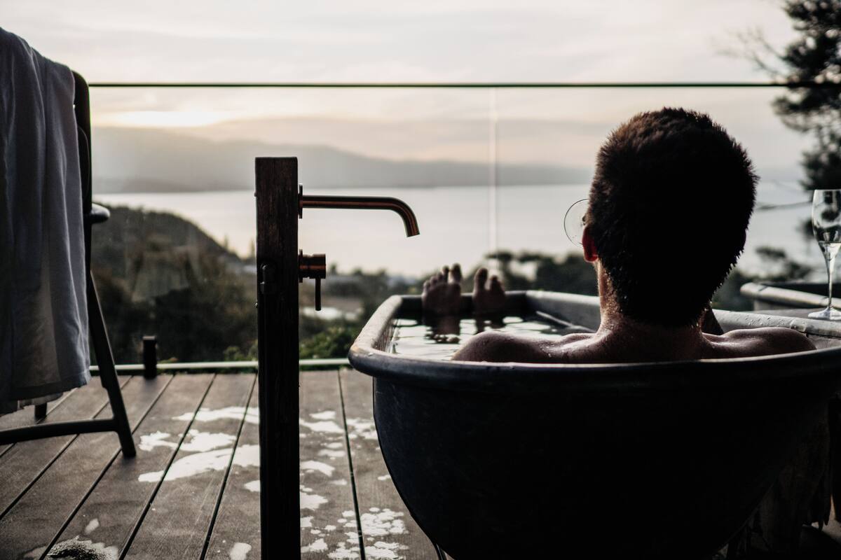 Uma pessoa em uma banheira com vista para um corpo d'água e colinas ao fundo.