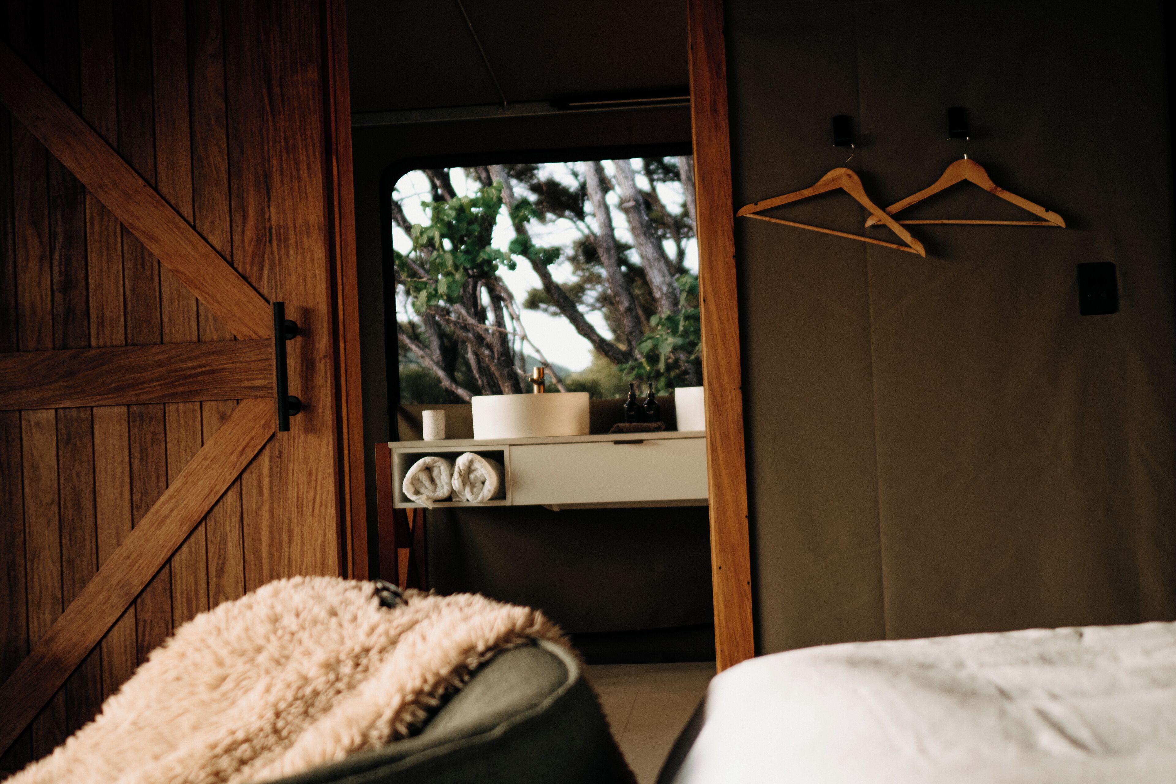 Quarto com porta de madeira, uma cama, uma mesinha com uma tigela e uma garrafa, e uma janela com vista para árvores.