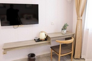 Quarto pequeno com televisão na parede, prateleira de madeira com chaleira e copos, uma cadeira e planta em vaso.