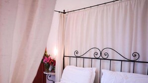 Uma cama bem arrumada com lençóis e almofadas brancas, uma cabeceira de metal preto e uma cortina branca.