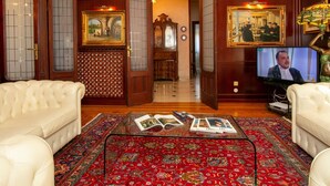 Uma sala de estar tradicional com um tapete vermelho estampado, dois sofás de couro branco, uma televisão e diversas obras de arte emolduradas nas paredes.