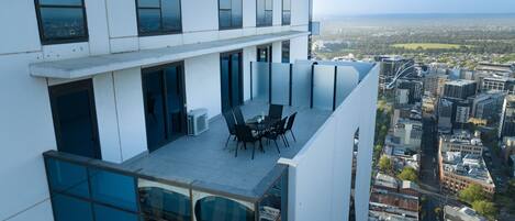 Um edifício moderno com terraço no telhado, apresentando uma mesa, cadeiras, grandes janelas e vista para a paisagem urbana.