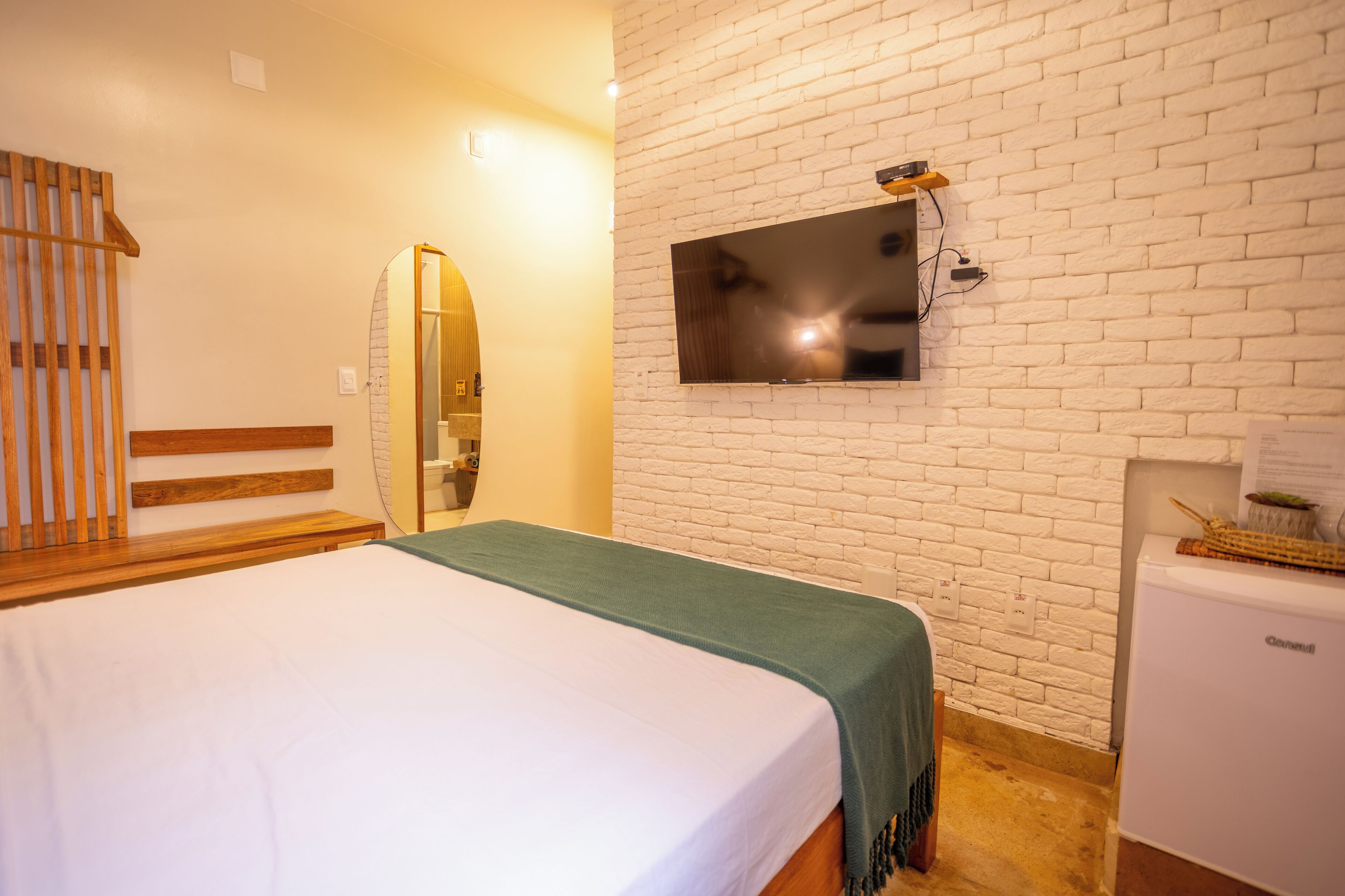 Quarto com cama, televisão fixada em parede de tijolos, pequena geladeira e um banco de madeira.