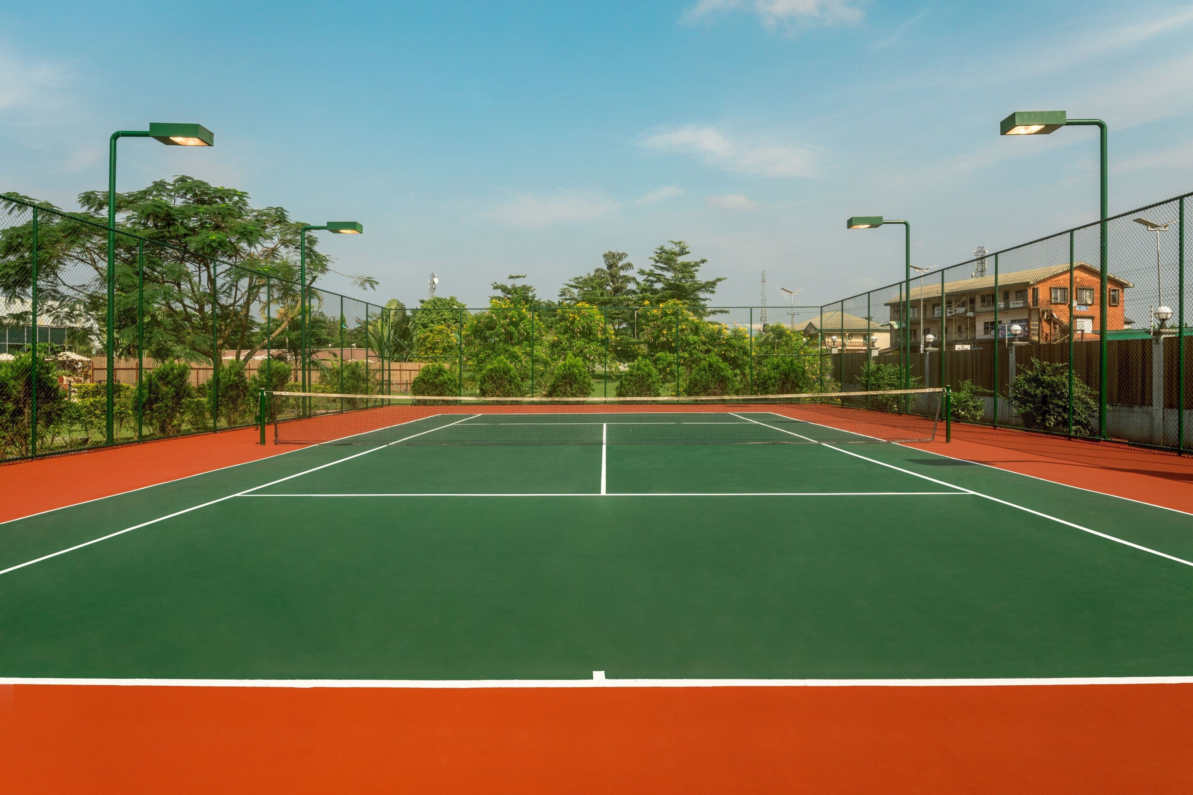 Uma quadra de tênis com superfícies verde e vermelha, cercada por uma grade e árvores.