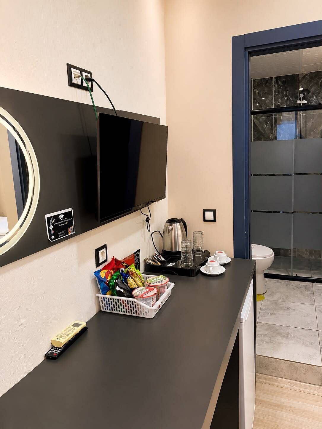 Quarto de hotel moderno com televisão de tela plana na parede, cafeteira, cesta de lanches, controle remoto e box de vidro.