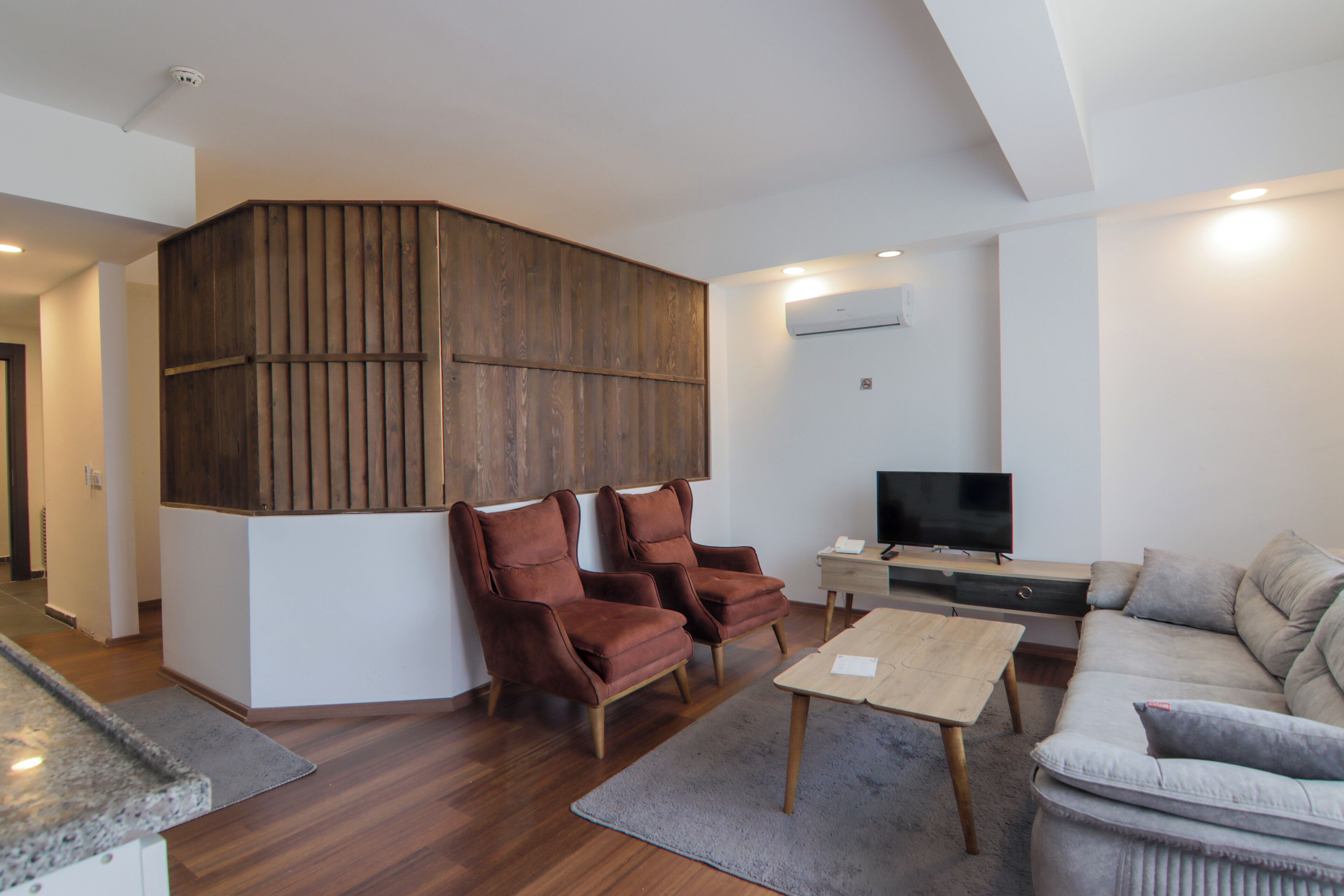 Uma sala moderna com uma parede de destaque em madeira, duas poltronas marrons, um sofá cinza, uma mesa de centro e uma televisão de tela plana.