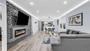 Uma sala de estar moderna com lareira, sofá cinzento e área de jantar ao fundo.