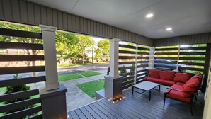 Um alpendre coberto com um sofá vermelho e uma mesa pequena, piso de madeira e vista para a rua e vegetação.