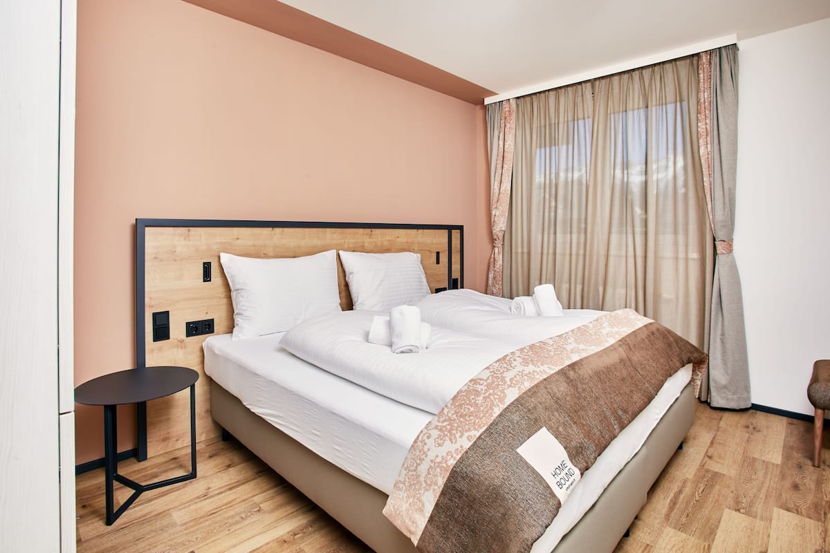 Uma cama bem arrumada com lençóis brancos e um cobertor marrom decorativo, uma pequena mesa redonda e uma cadeira em um quarto com piso de madeira e paredes cor de pêssego.