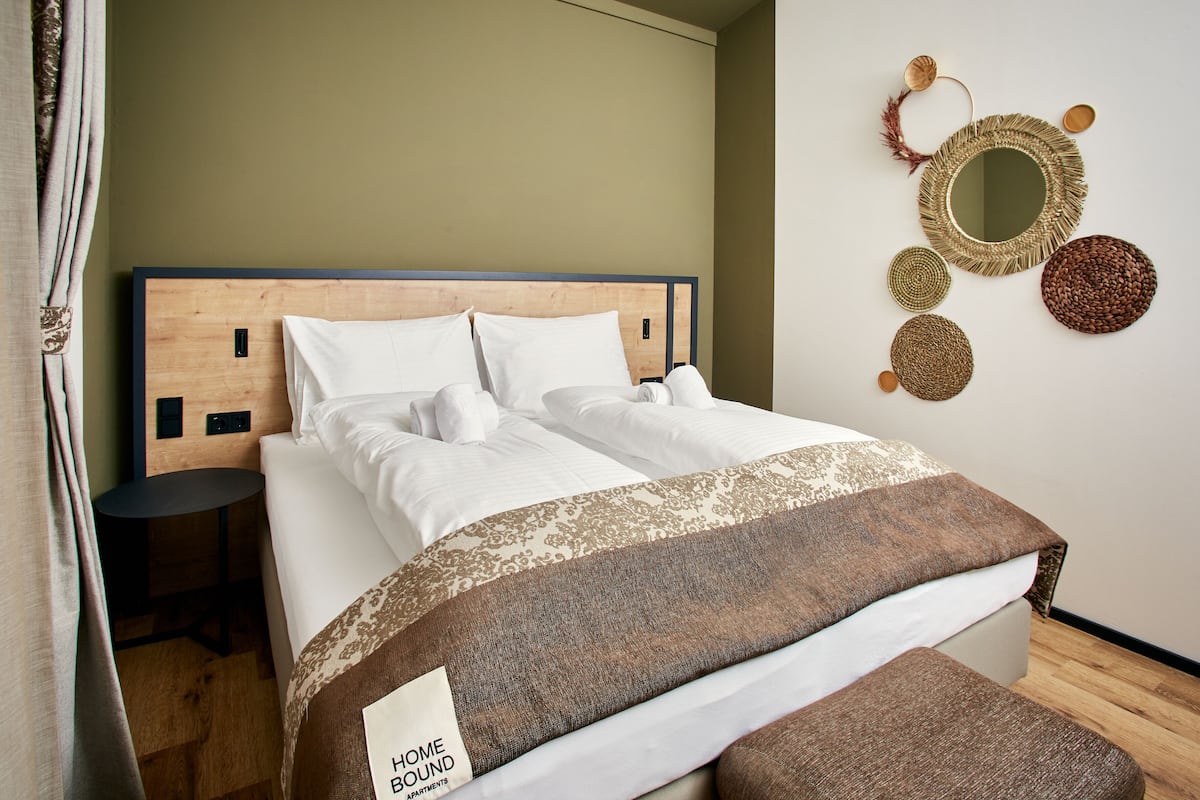 Cama bem arrumada com colcha marrom e branca estampada, uma pequena mesa de cabeceira e um espelho fixado na parede com objetos decorativos.