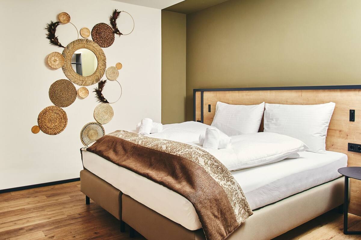 Quarto de hotel moderno com cama, cabeceira, espelho fixado na parede e arte decorativa em tecido.