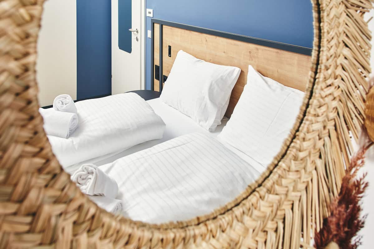 Cama com lençóis brancos e cabeceira de madeira, cesta de palha com toalhas, e parede azul ao fundo.